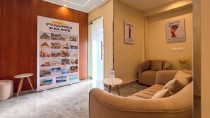 Um hall de hotel com uma mesa de recepção, uma área de estar com dois sofás, uma pequena mesa com uma garrafa de água e um grande cartaz informativo sobre as Pirâmides de Gizé.