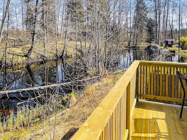 Um deck de madeira com vista para uma área arborizada, um pequeno lago e uma cabana.