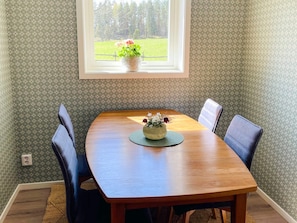 Área de refeições com mesa de madeira, quatro cadeiras, um vaso com flores e uma janela com vista para árvores.