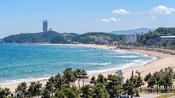 Uma praia costeira com areal, mar azul límpido e um cenário de edifícios e colinas.