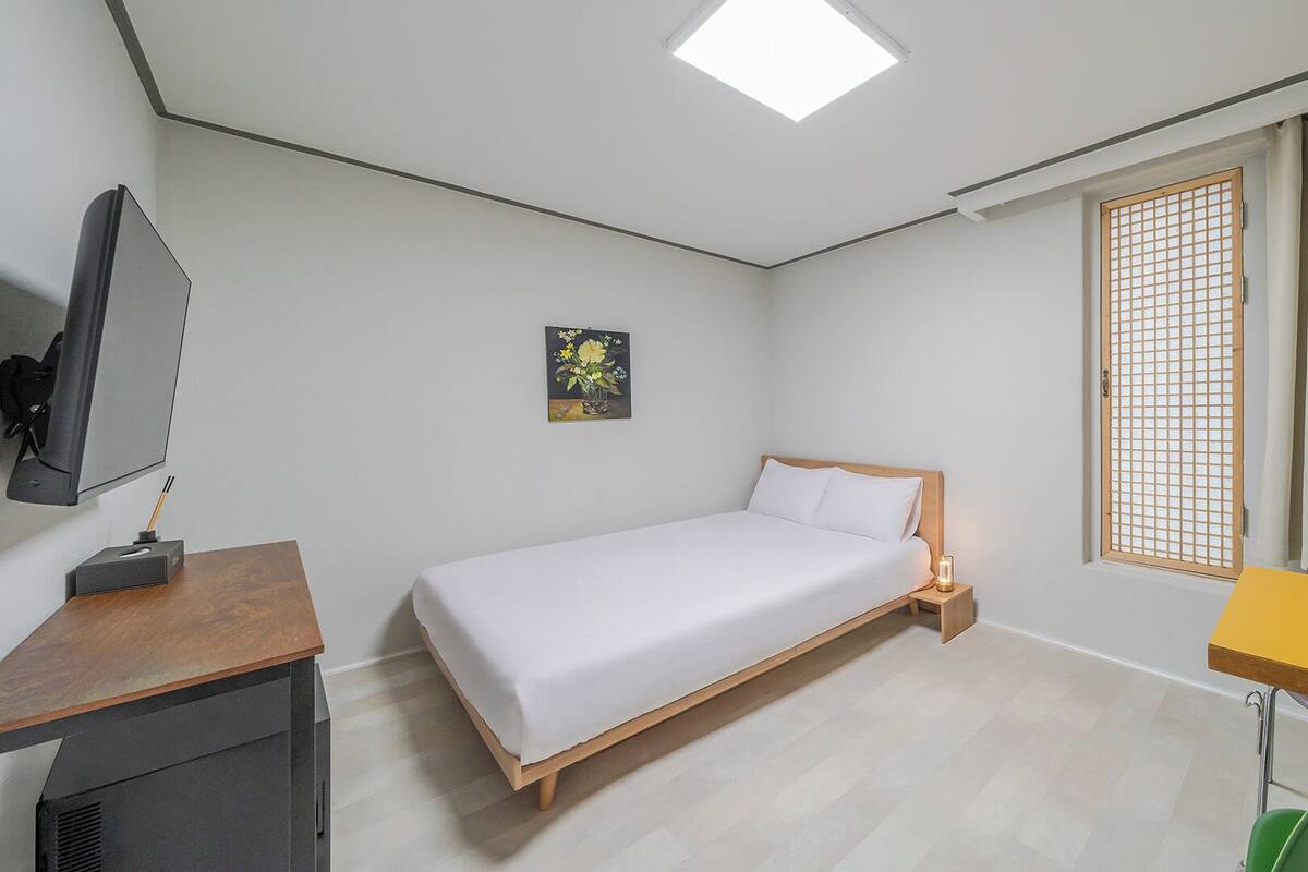 Um quarto com cama, escrivaninha, televisão, janela com cortinas e um quadro na parede.