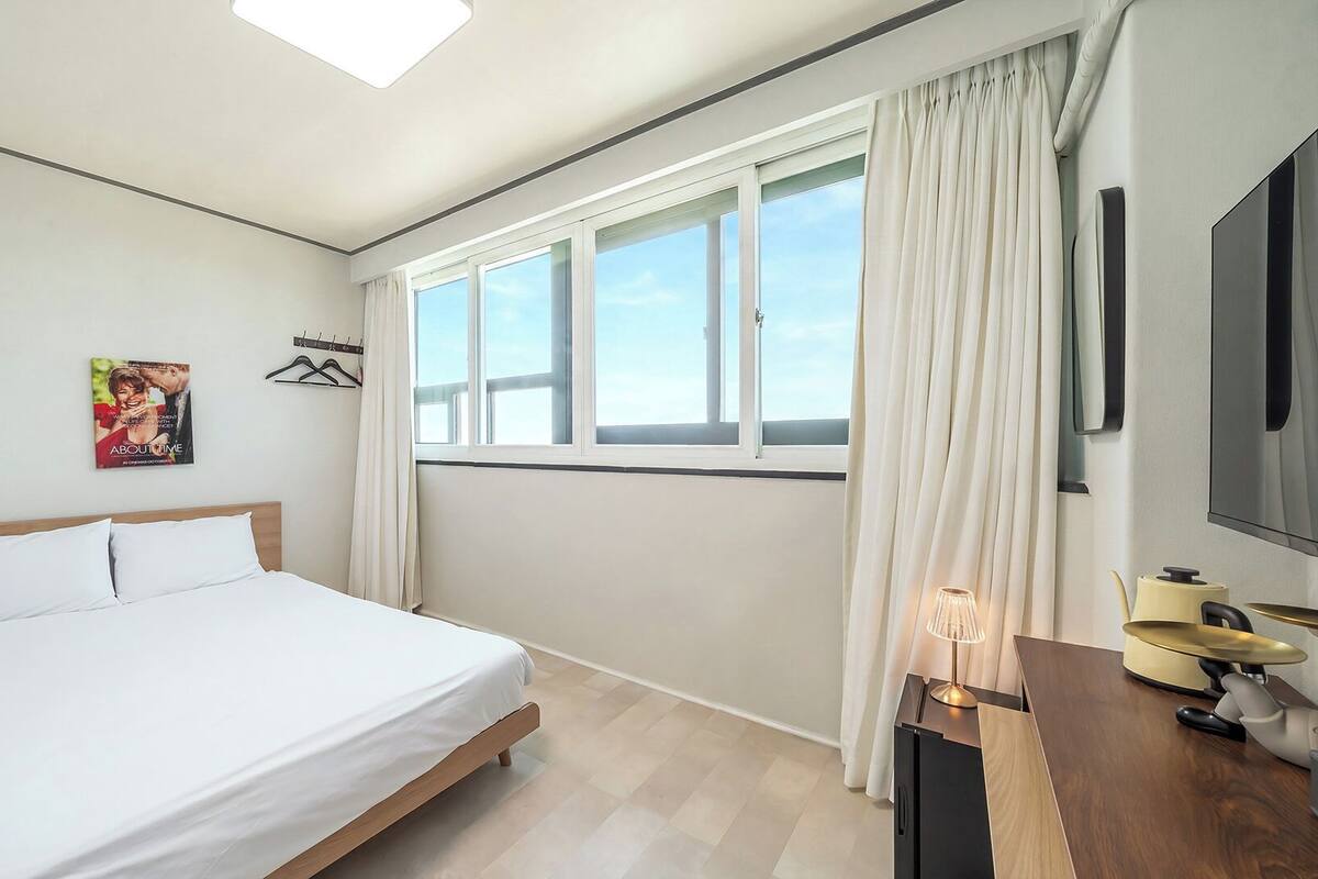 Quarto de hotel com cama, televisão, secretária com candeeiro e janela com cortinas.