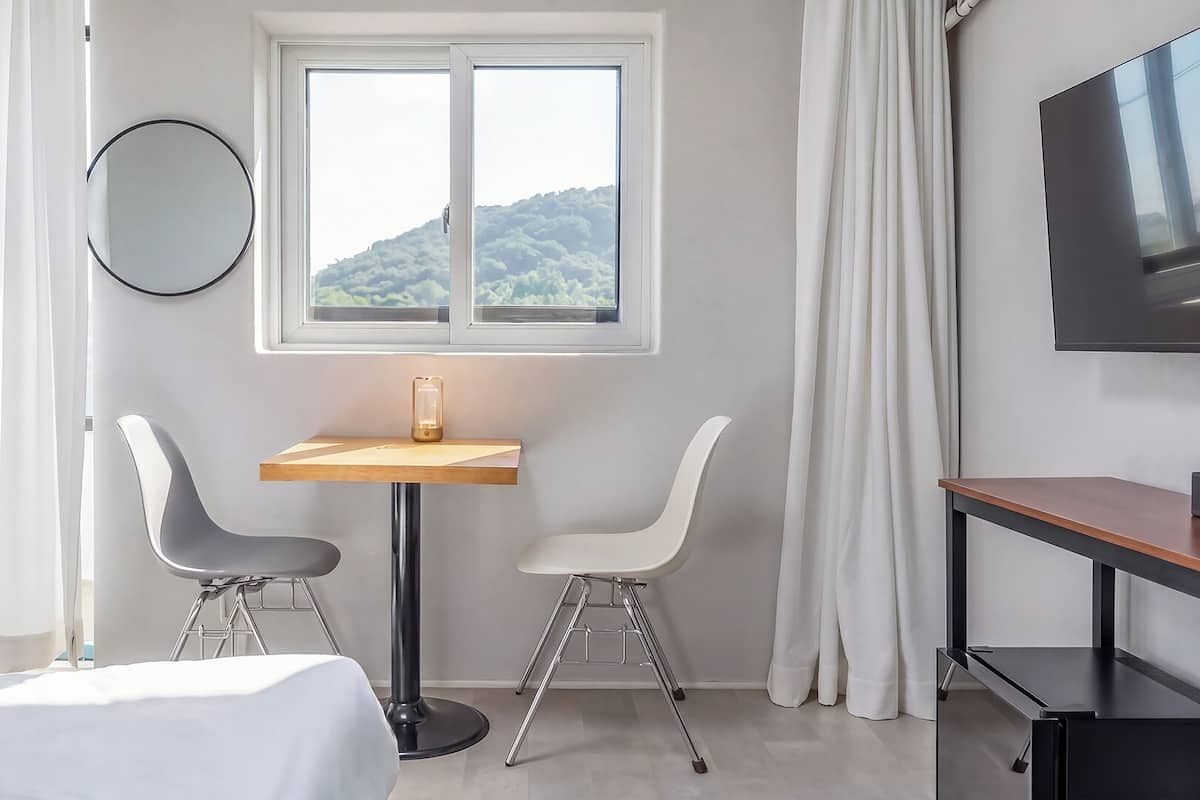 Quarto de hotel com uma cama, uma mesa com duas cadeiras, uma televisão, uma janela com vista para áreas verdes e um espelho redondo na parede.
