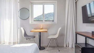Quarto de hotel com uma cama, uma mesa com duas cadeiras, uma televisão, uma janela com vista para áreas verdes e um espelho redondo na parede.