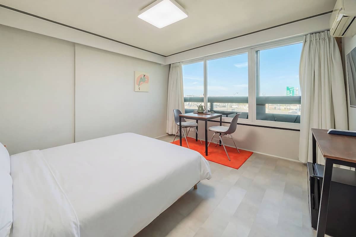 Quarto de hotel com cama, escrivaninha, cadeira e janela com vista.