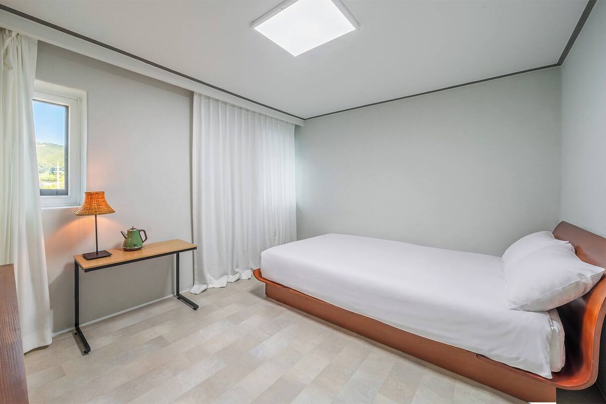 Quarto com cama, mesa de cabeceira com abajur, janela com cortinas e luminária no teto.