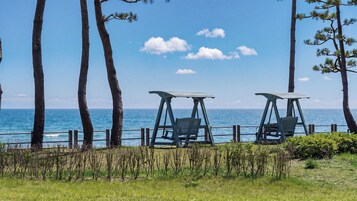 Vista costeira com duas cadeiras de baloiço e uma mesa, rodeada por árvores altas e uma vedação de madeira.
