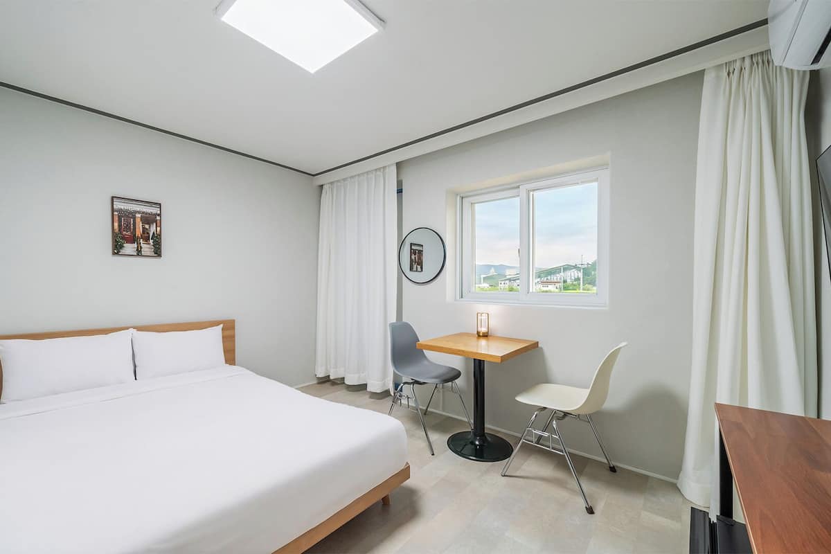 Quarto de hotel com cama, escrivaninha, duas cadeiras, janela com cortinas e um quadro na parede.