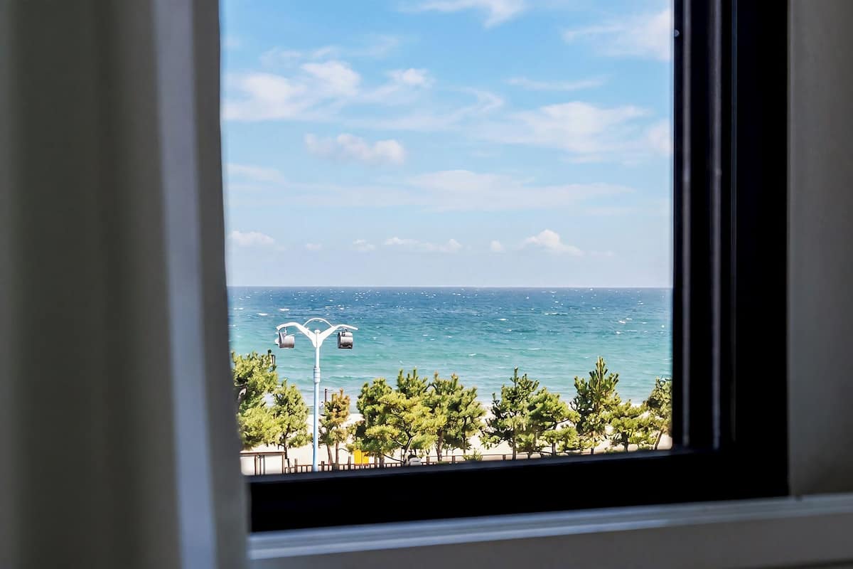 Vista pela janela de uma área costeira com um lampião, árvores e o oceano.