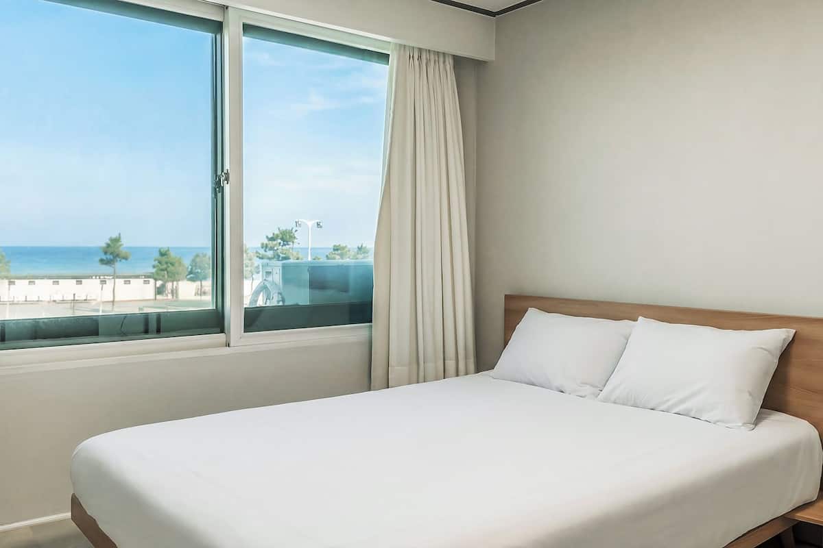Quarto de hotel com cama, janela e vista para a praia, e cortina.