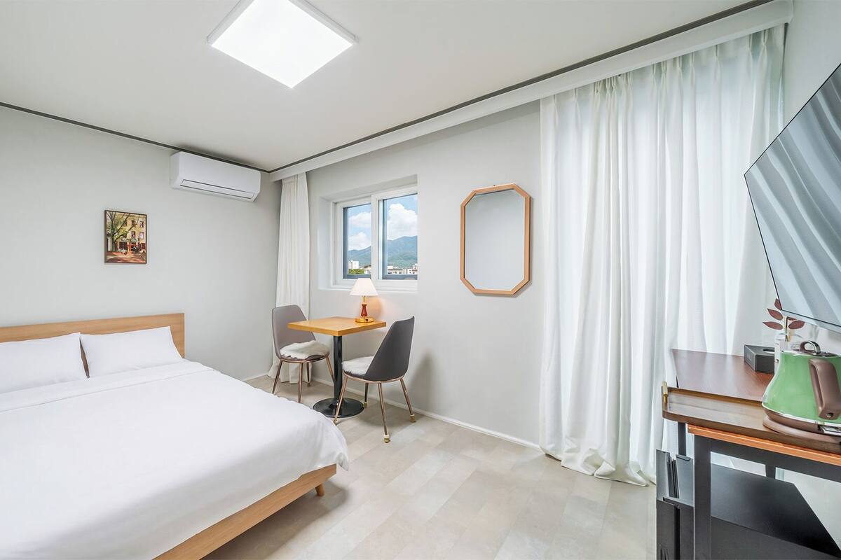 Quarto de hotel com cama, escrivaninha, cadeira, janela com cortinas, aparelho de ar condicionado e um quadro na parede.