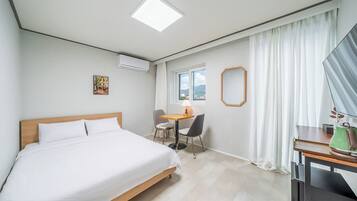 Quarto de hotel com cama, escrivaninha, cadeira, janela com cortinas, aparelho de ar condicionado e um quadro na parede.