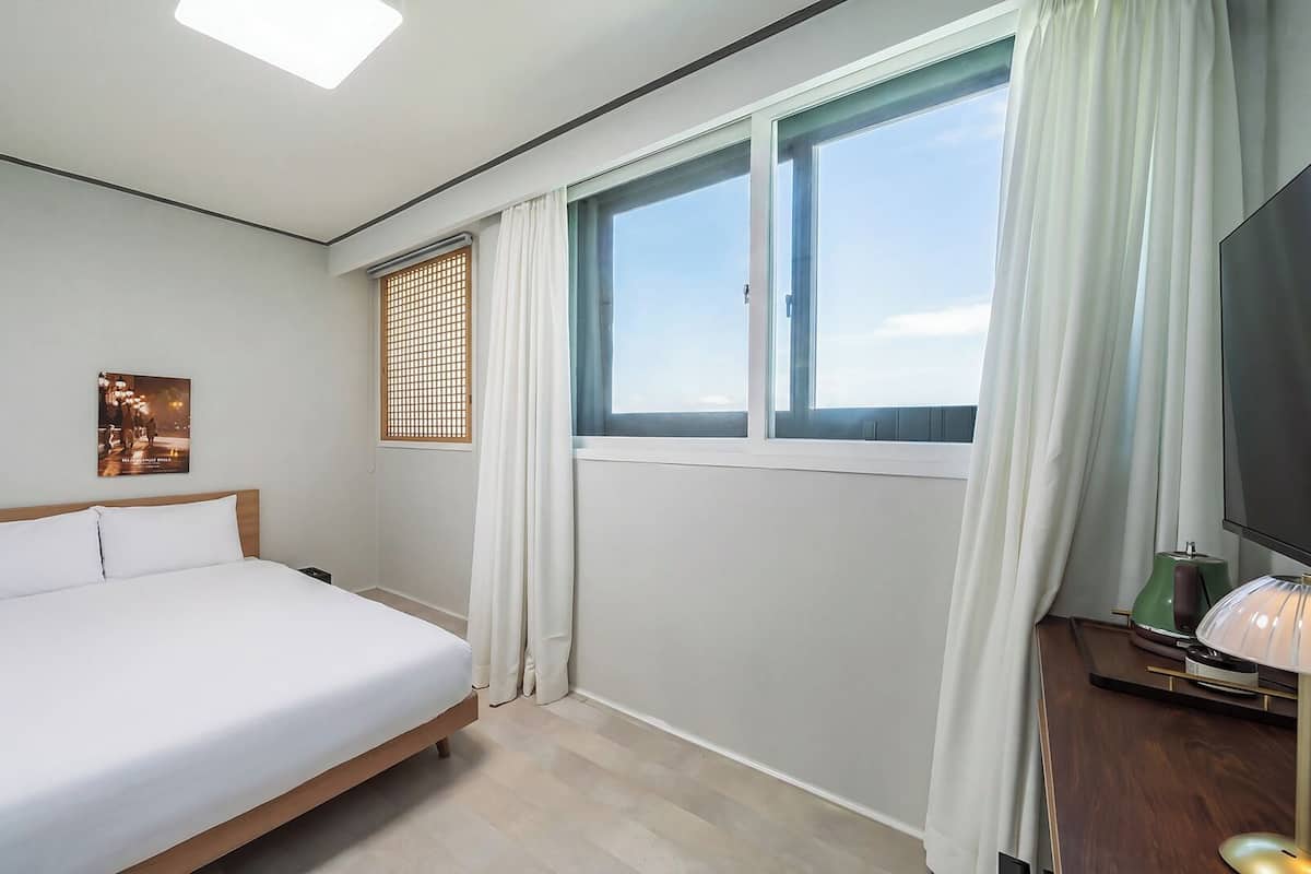 Quarto de hotel com cama, janela com cortinas, televisão montada na parede e uma mesinha com abajur.