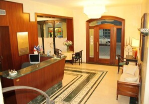 Um hall de hotel com balcão de recepção, cadeiras, uma pequena mesa com um vaso de flores e uma porta que dá para o exterior.