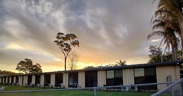Uma fileira de edifícios térreos com grandes janelas, possivelmente um hotel ou hostel, com uma área gramada na frente e árvores ao fundo.