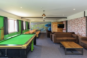 Uma mesa de bilhar, sofás, uma TV e um mural em um quarto com paredes verdes e teto de madeira.