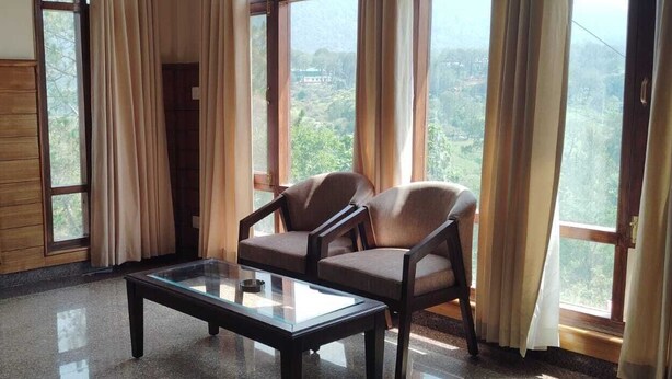 Quarto com mesa de vidro, duas cadeiras marrons, janelas amplas com cortinas e vista para áreas verdes.