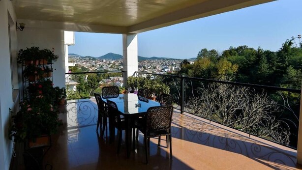 Uma área de refeições ao ar livre coberta, com mesa e cadeiras, plantas em vasos e vista para um horizonte urbano e montanhas.