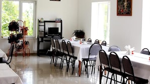Uma sala de jantar com mesas e cadeiras, uma estante de livros e uma janela com vista para o verde.