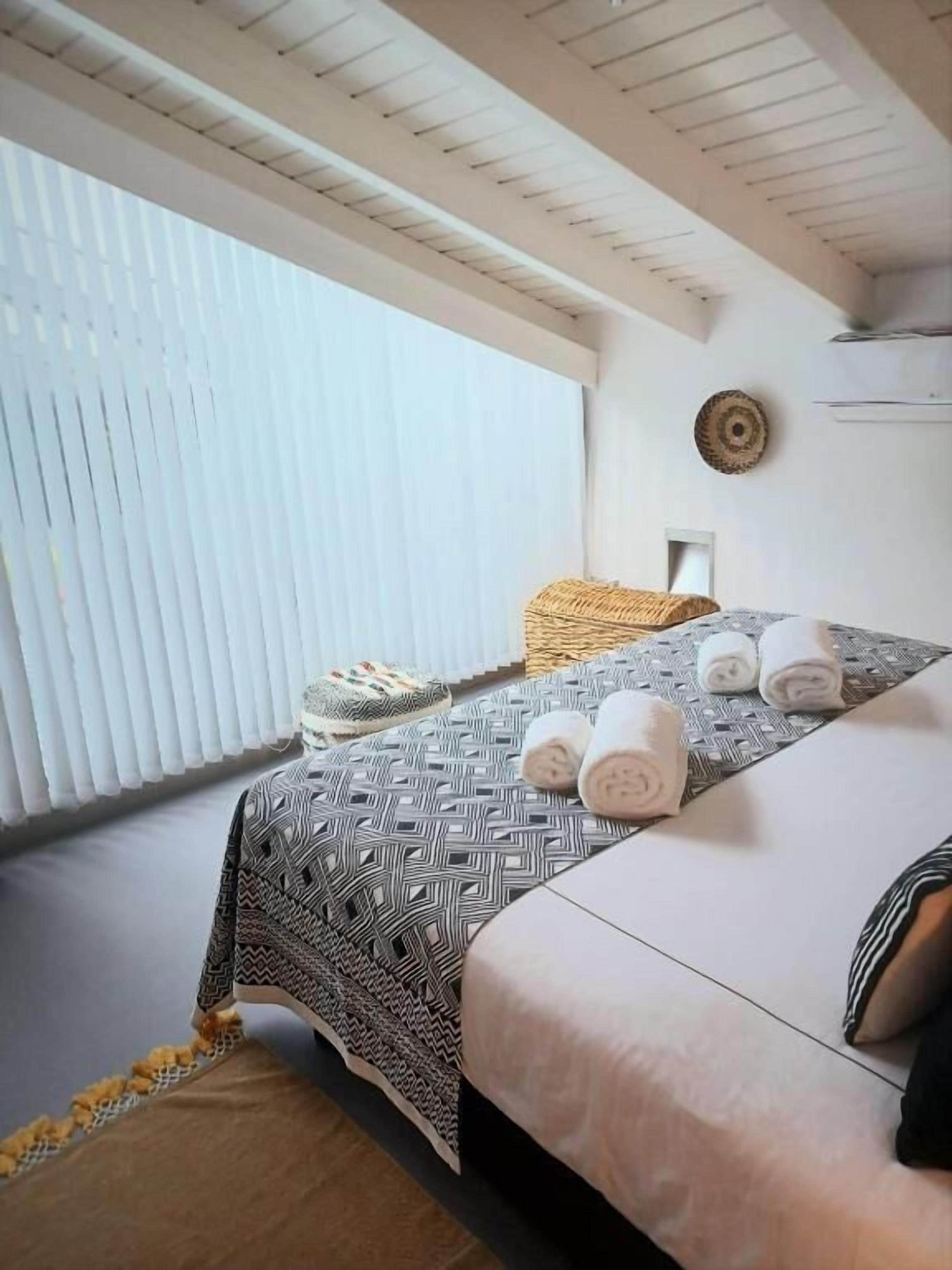 Quarto com cama, janela com persianas, ar-condicionado fixado na parede e cesta de fibra natural no chão.