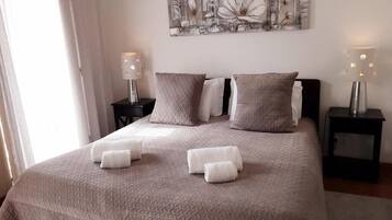 Cama bem arrumada com colcha texturizada, dois travesseiros e duas toalhas enroladas. Há uma obra de arte na parede acima da cama e duas luminárias de cabeceira de cada lado.
