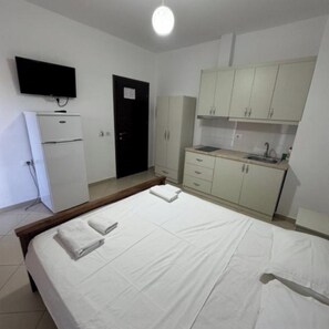 Um quarto com cama, televisão fixada na parede, porta e cozinha compacta com armários.
