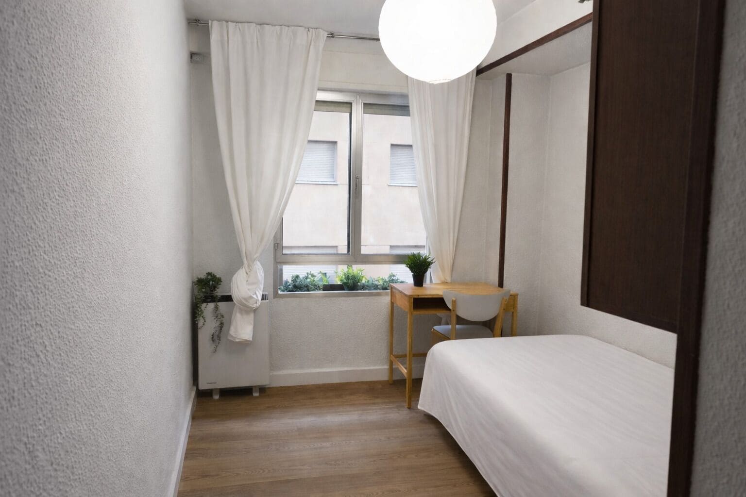 Quarto pequeno e bem arrumado, com cama, escrivaninha e janela com cortinas.