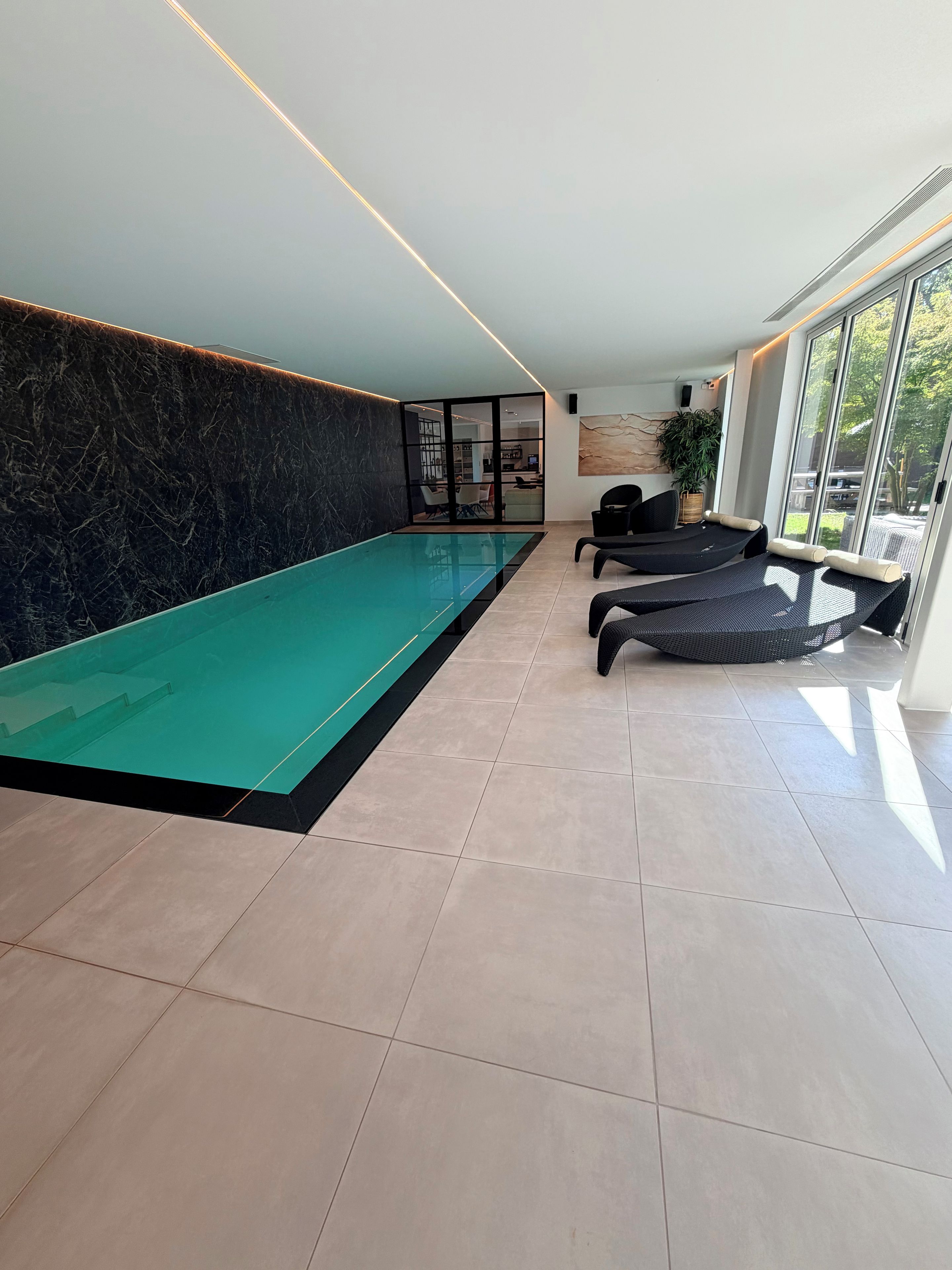 Piscina interior moderna com fundo de vidro, ao lado de uma área de estar com espreguiçadeiras e uma grande janela com vista para o exterior.