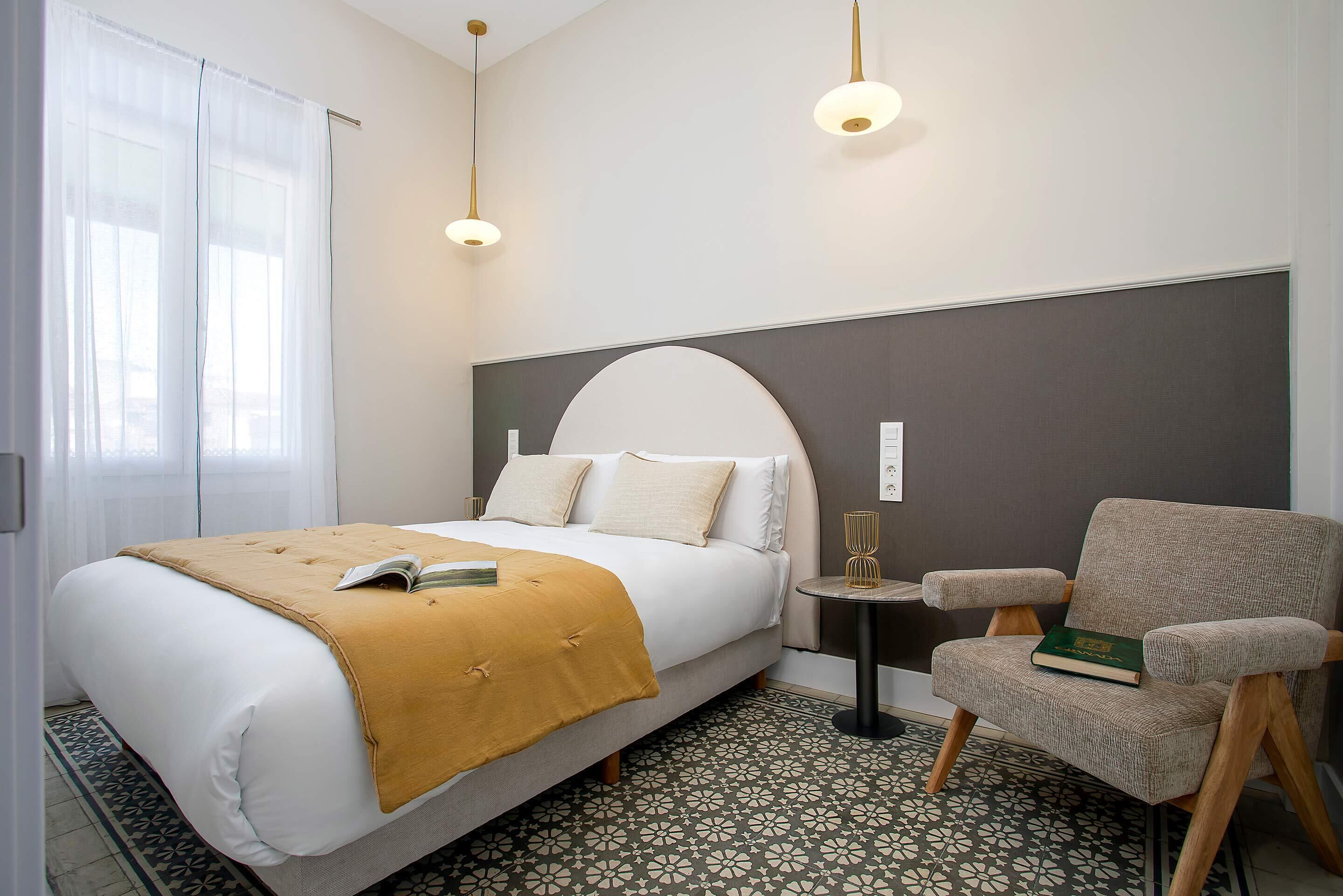 Quarto de hotel com cama, uma cadeira, uma mesinha e chão estampado.