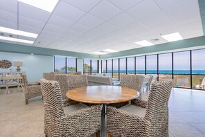 Um quarto amplo com uma mesa redonda e cadeiras de vime, janelas grandes com vista para o mar e um teto com iluminação embutida.