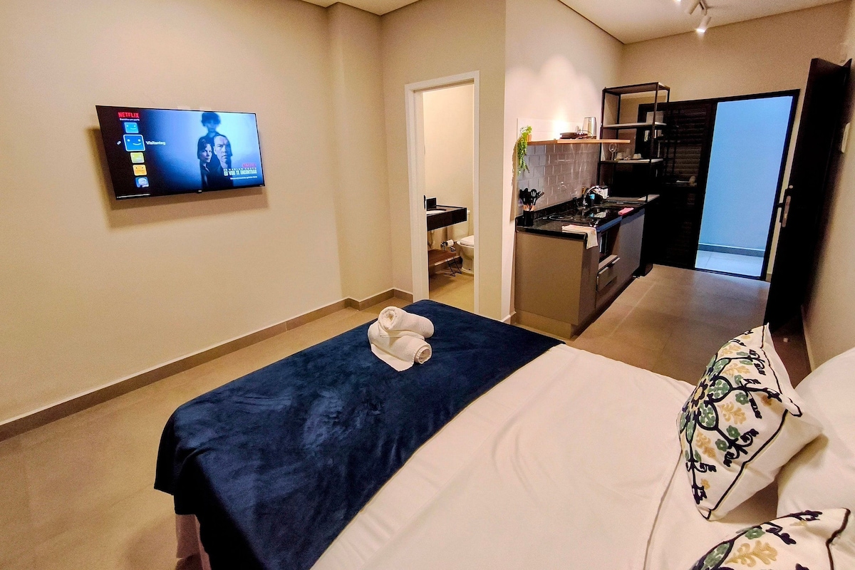 Quarto de hotel com cama, televisão presa na parede, cozinha compacta e banheiro visível por uma porta aberta.