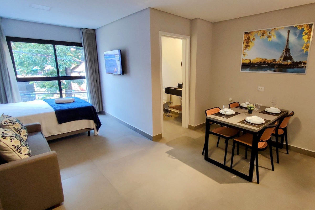 Quarto de hotel com cama, mesa de jantar posta para dois, pintura da Torre Eiffel e janela com vista para o verde.
