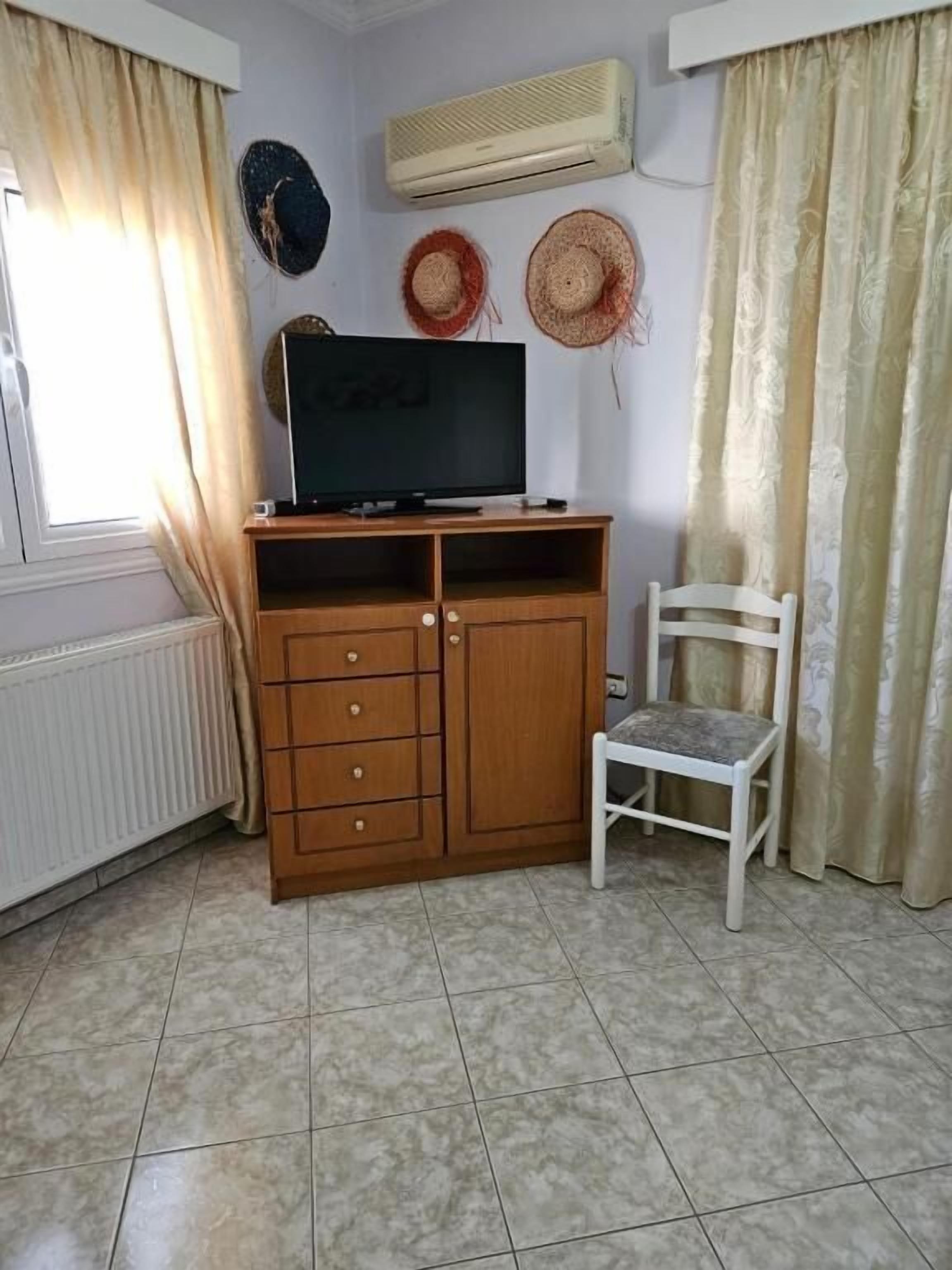Quarto com televisão sobre um armário de madeira, uma cadeira branca e um radiador.