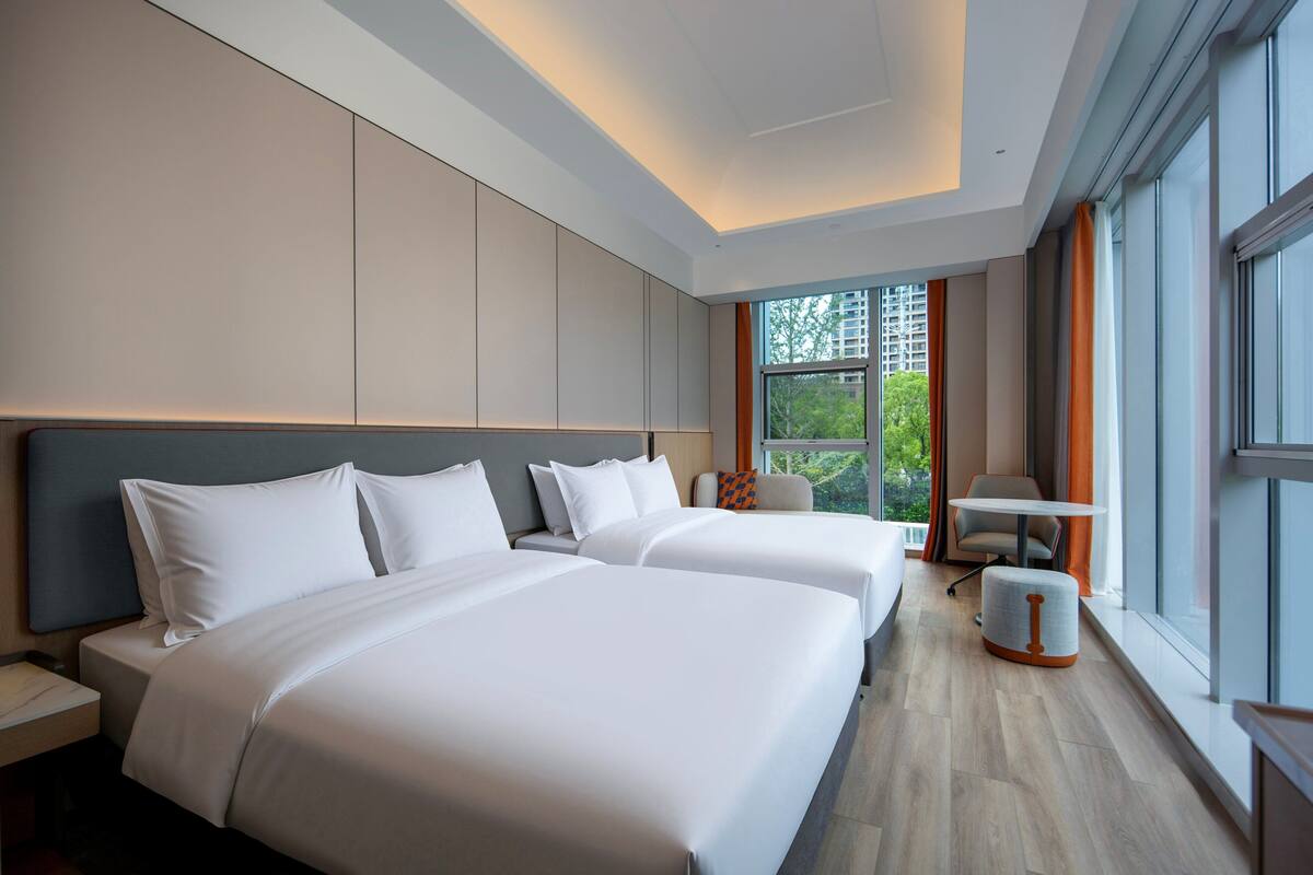 Quarto de hotel moderno com duas camas, uma mesinha e uma janela com vista para a cidade.