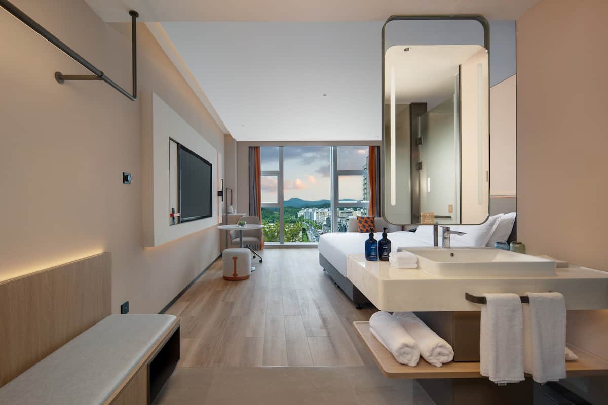 Quarto de hotel moderno com uma cama grande, banheiro com um espelho amplo, um banco e vista para a cidade.