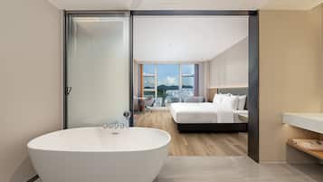 Quarto moderno com uma banheira grande, cama com muitos travesseiros e vista da cidade através de amplas janelas.