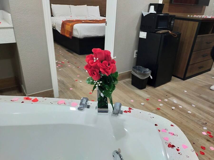 Quarto de hotel com uma banheira branca grande, uma cama com cobertor branco e laranja, um cesto de lixo preto pequeno e um micro-ondas em um armário de madeira.