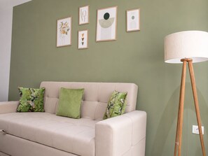 Um sofá bege com almofadas florais verdes, uma luminária de chão e quatro quadros emoldurados numa parede verde.