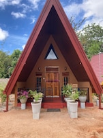 Um edifício triangular com porta de madeira, plantas em vasos e telhado vermelho.