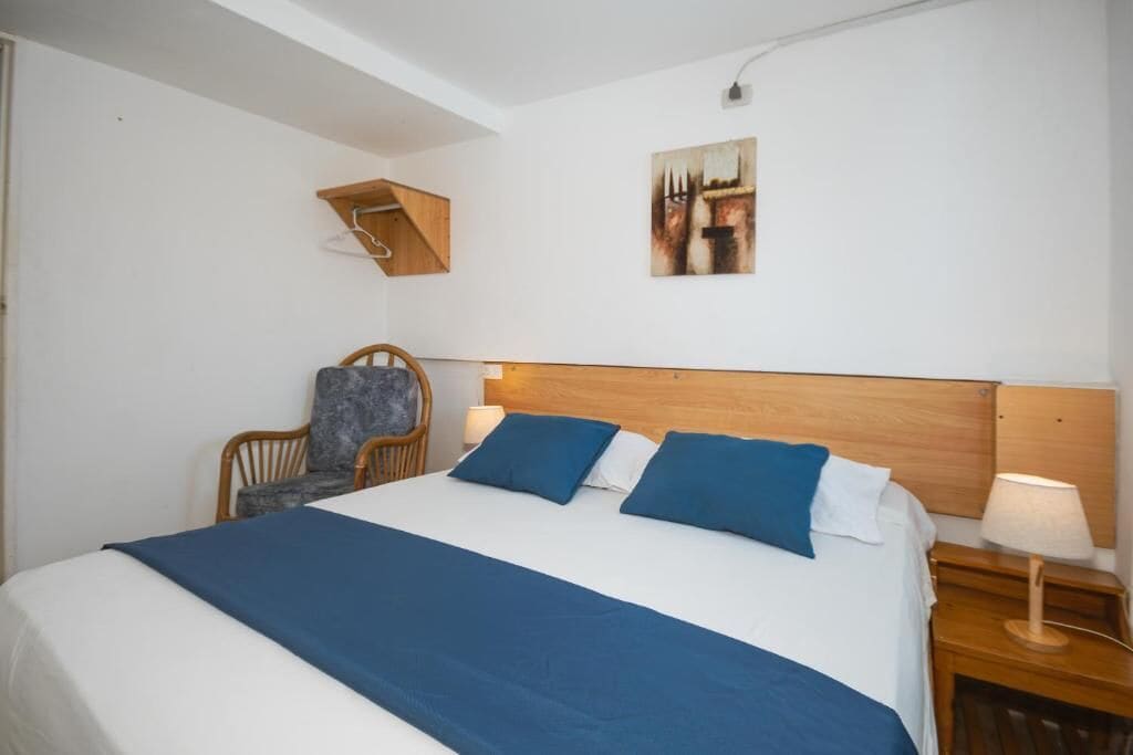 Uma cama com cabeceira de madeira, edredom azul e duas almofadas. Há uma mesa de cabeceira de madeira com uma luminária, uma cadeira e uma prateleira fixada na parede.