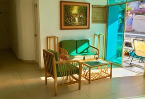 Quarto com sofá verde, cadeiras de madeira, uma mesa pequena e um quadro na parede.
