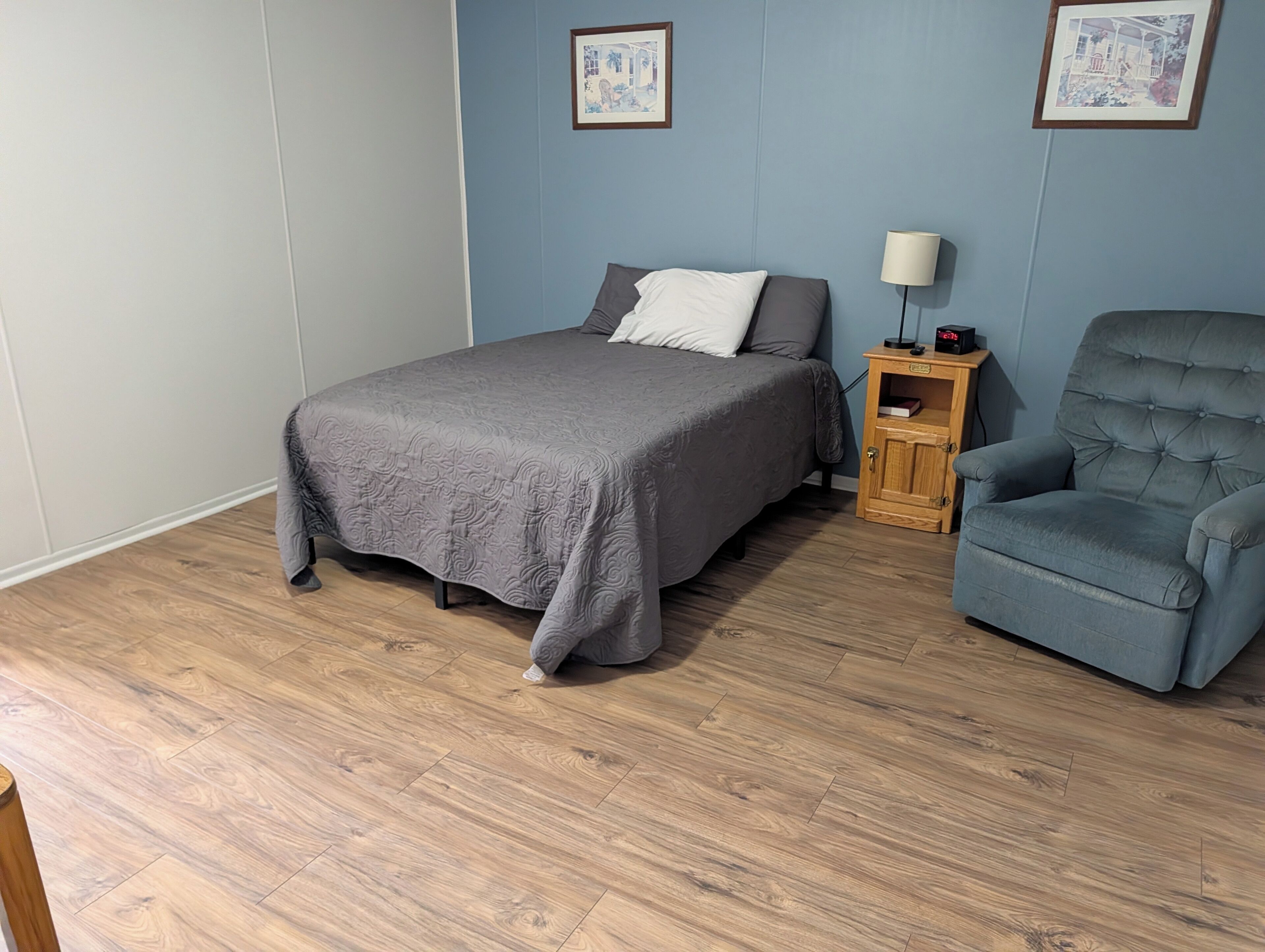 Quarto com cama, uma cadeira, mesa de cabeceira e duas imagens emolduradas na parede.