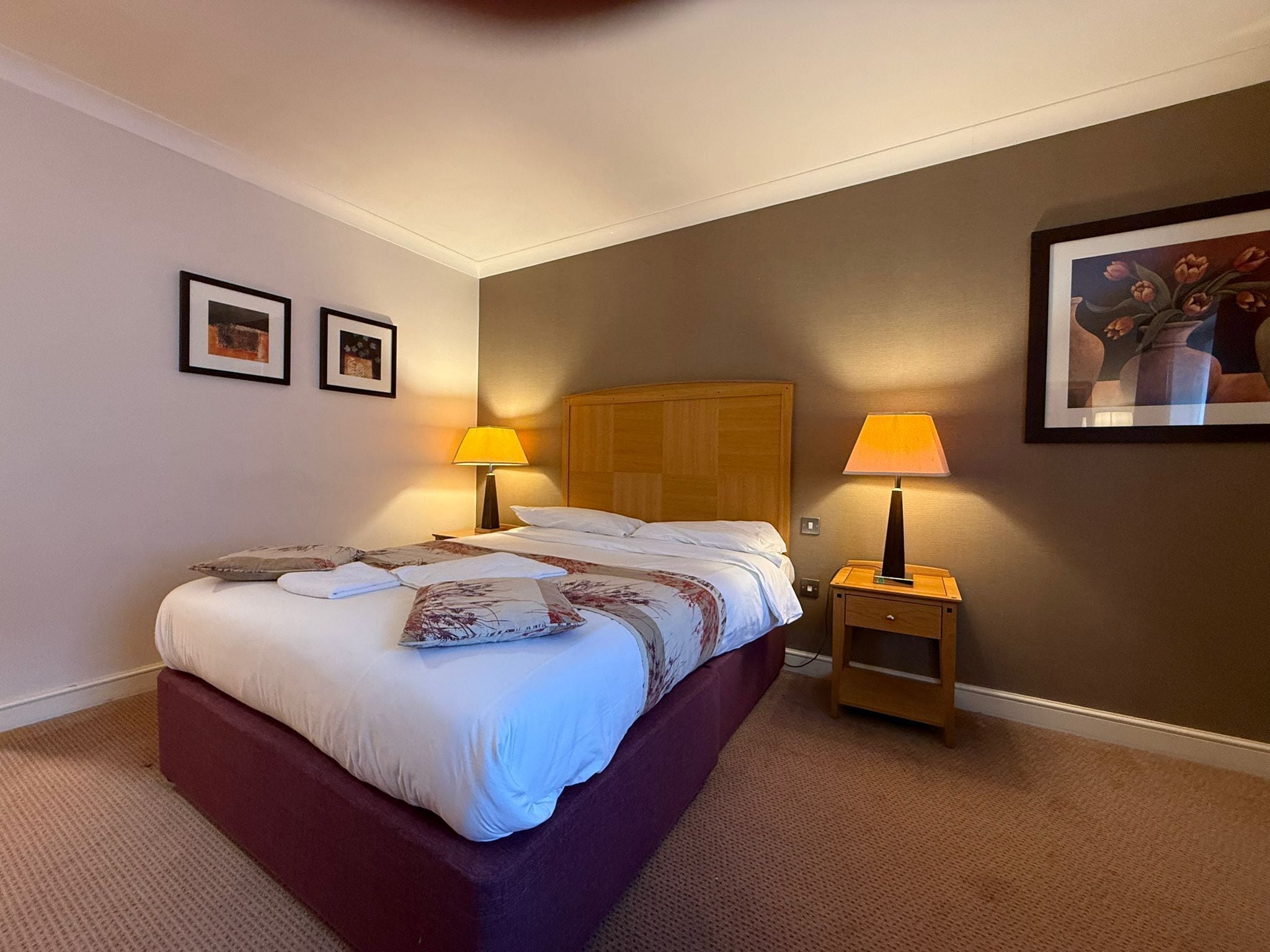 Quarto de hotel com uma cama, duas mesas de cabeceira com abajures, uma cabeceira de madeira e duas imagens emolduradas na parede.