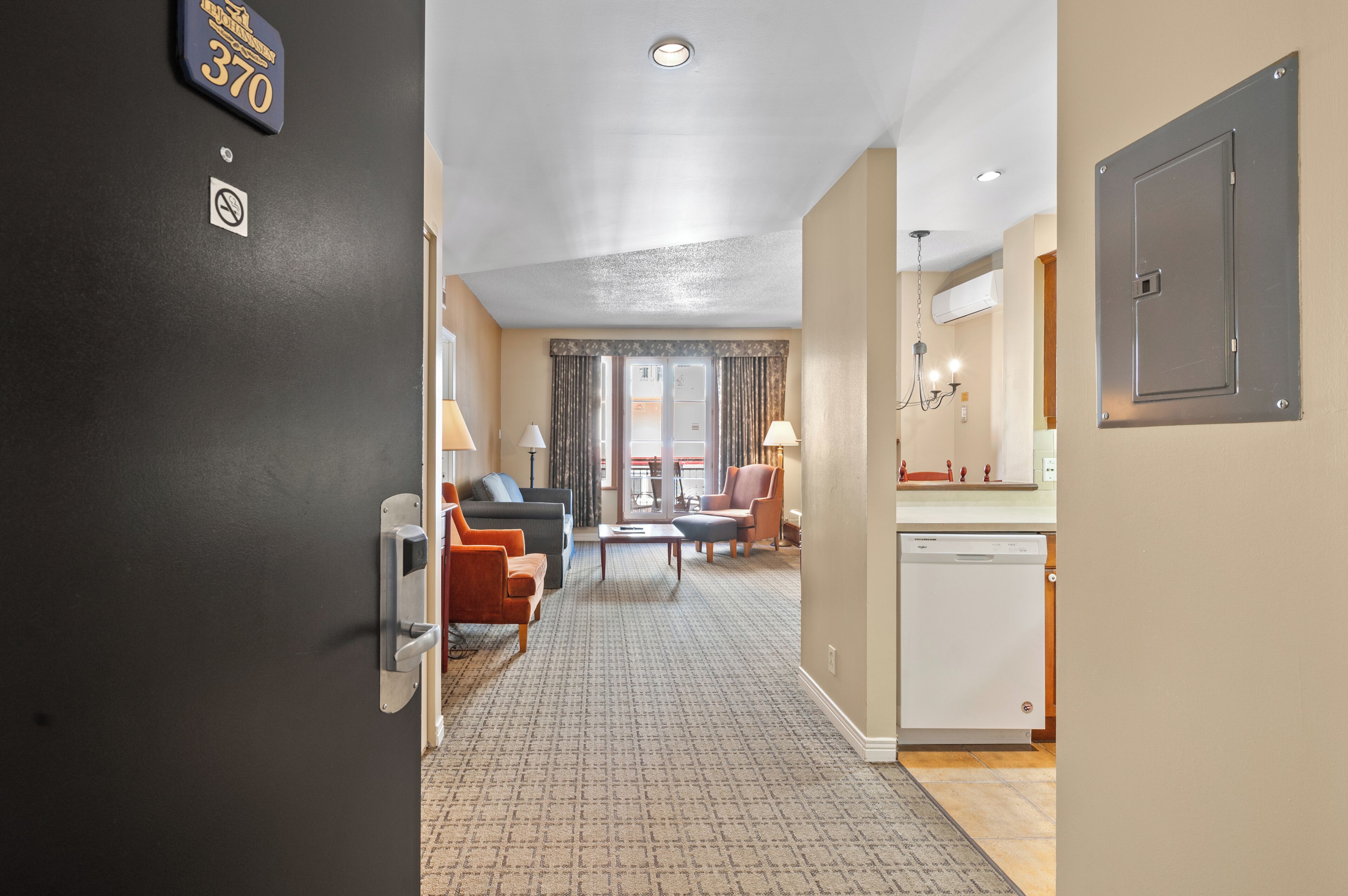 Quarto de hotel com porta escura, placa de número, placa de proibido fumar, sala de estar com sofá, cadeiras, mesa de centro, cozinha com frigorífico e janela com cortinas.