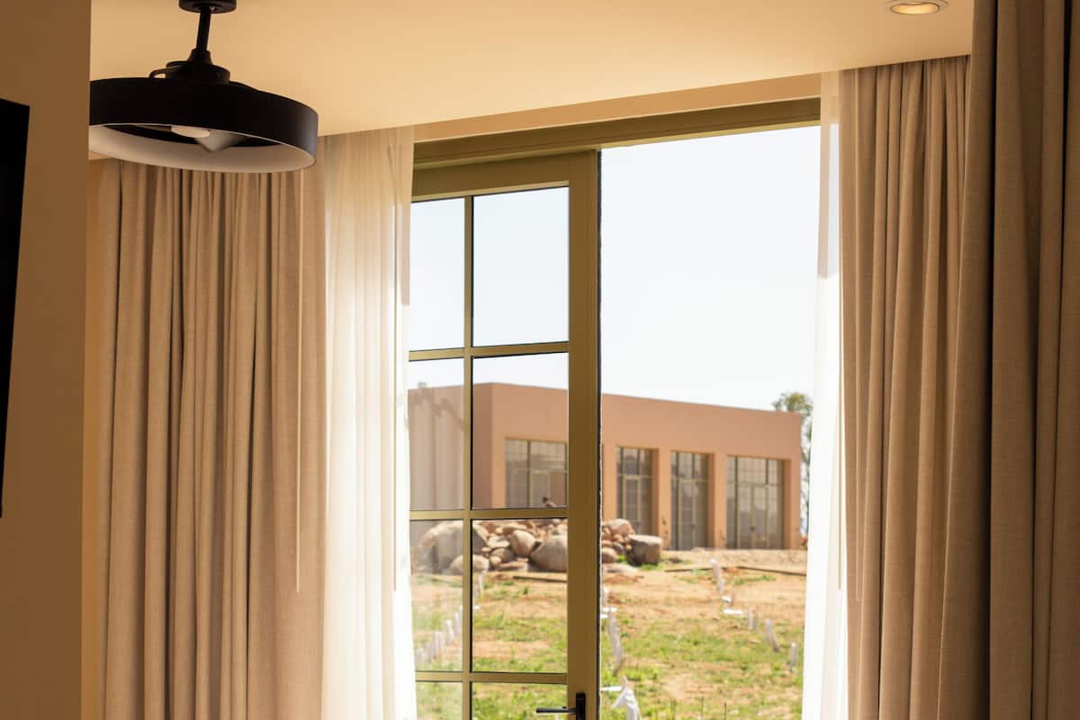 Quarto com cama, mesa de cabeceira de madeira e vista para um edifício e área externa através de uma porta de vidro.