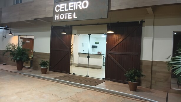 Entrada de hotel com uma grande placa indicando 'CELEIRO HOTEL', uma recepção e plantas em vasos.