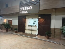 Entrada de hotel com uma grande placa indicando 'CELEIRO HOTEL', uma recepção e plantas em vasos.