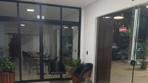 Um hall de hotel com balcão de recepção, portas de vidro e área de estar com cadeiras e plantas em vasos.