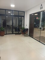 Um hall de hotel com balcão de recepção, portas de vidro e área de estar com cadeiras e plantas em vasos.