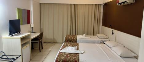 Quarto de hotel com duas camas, uma escrivaninha com computador, uma cadeira e ar condicionado.